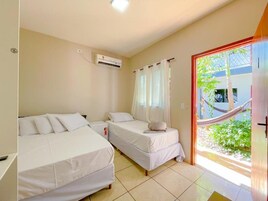 Quarto com duas camas, janela com cortinas, aparelho de ar condicionado e vista para o verde lá fora.