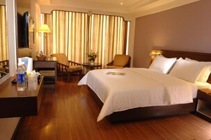 Quarto de hotel com uma cama grande, uma escrivaninha com luminária, uma cadeira, uma mesinha com um vaso e uma TV fixada na parede.