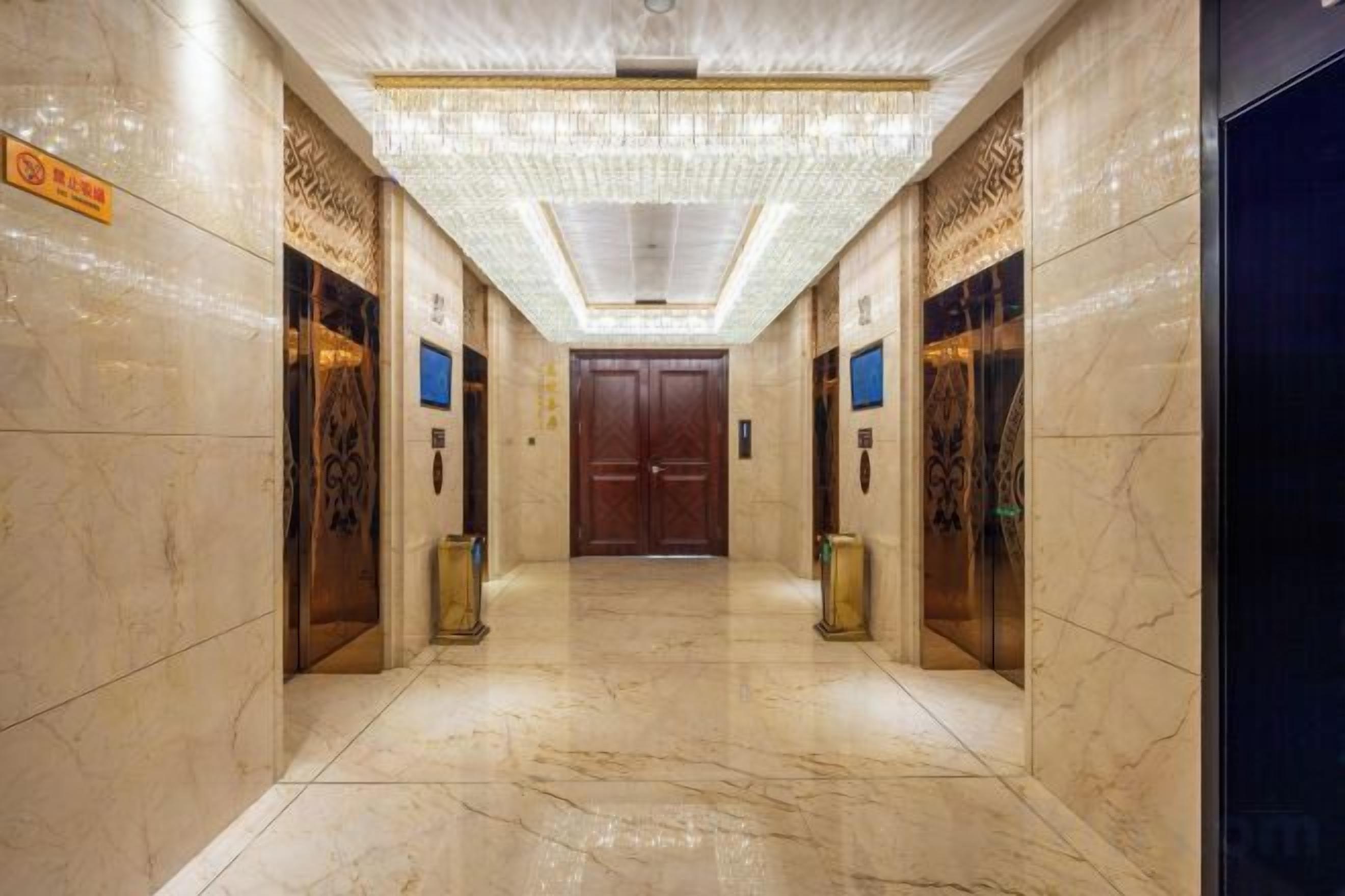 Um corredor com piso de mármore, um lustre e dois elevadores com portas.