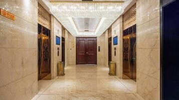 Um corredor com piso de mármore, um lustre e dois elevadores com portas.