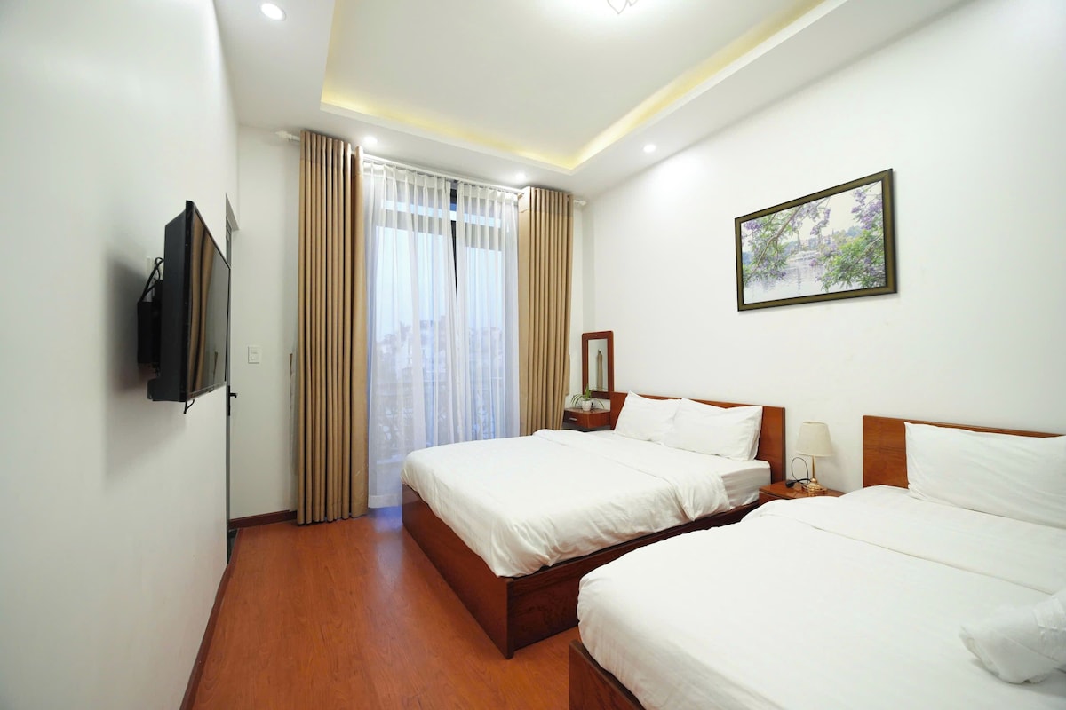 Quarto de hotel com duas camas, televisão, mesa de cabeceira com abajur e janela com cortinas.