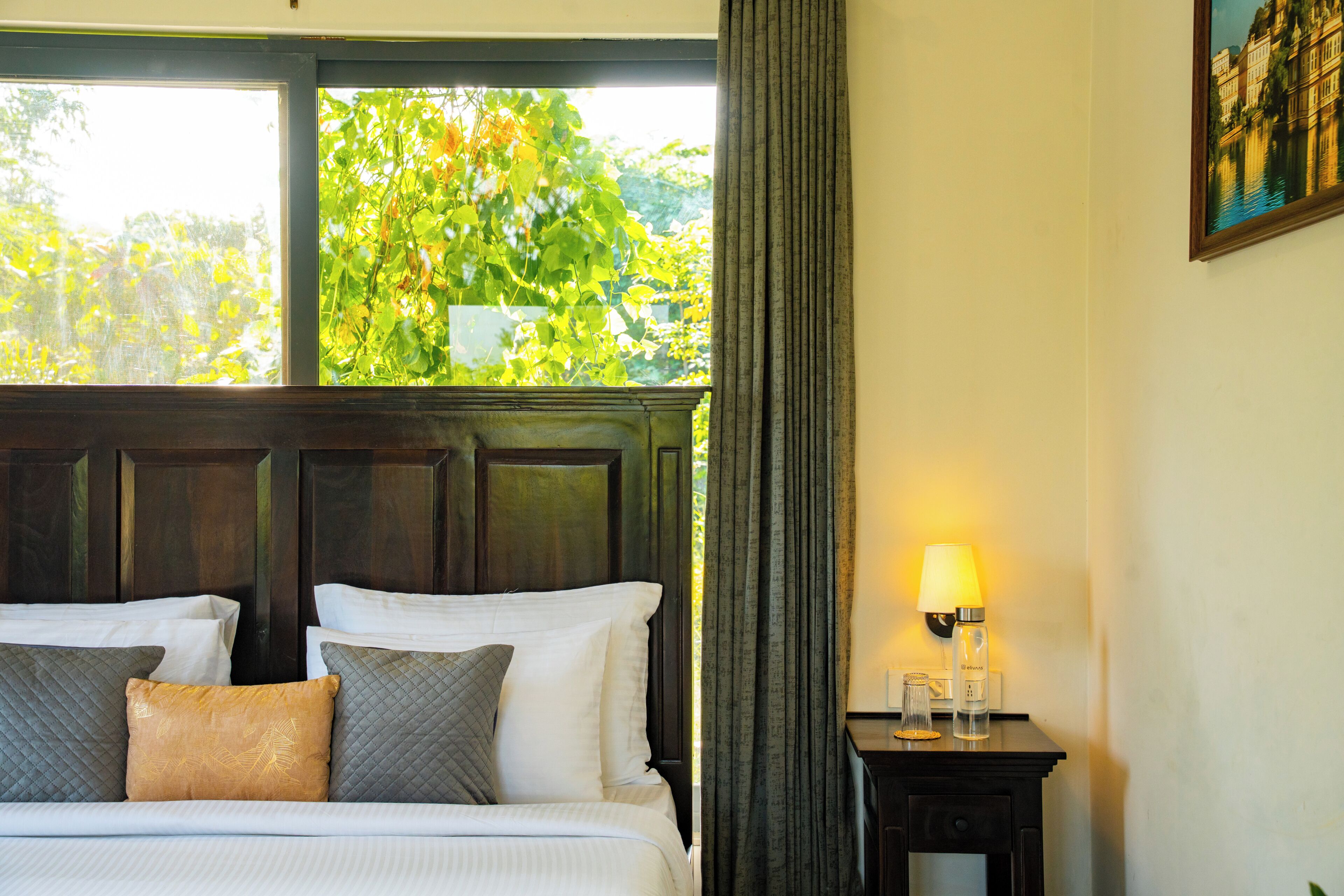 Quarto de hotel com uma cama grande, mesa de cabeceira com abajur e janela com vista para o verde.
