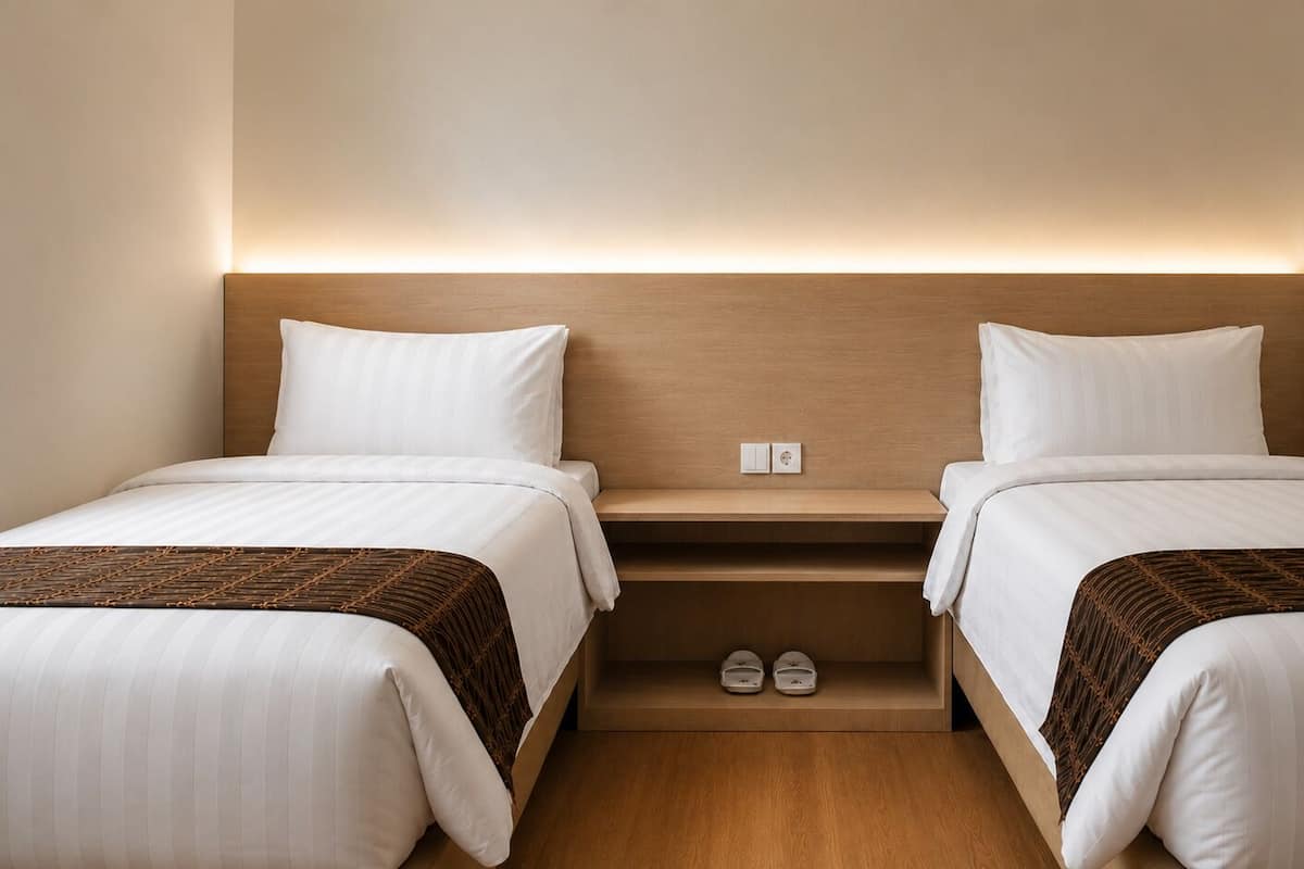 Quarto de hotel com duas camas, uma cabeceira de madeira, uma prateleira com sapatos e uma tomada na parede.