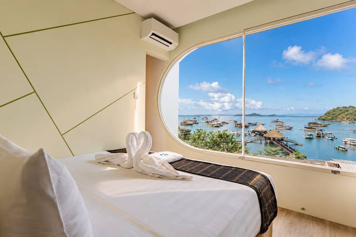 Quarto de hotel com cama, travesseiros, janela com vista para barcos e um cais, e um aparelho de ar condicionado.