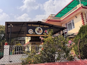 Uma área externa coberta com uma placa indicando culinária local, um jardim cercado com plantas floridas e um edifício com telhado vermelho e paredes bege.