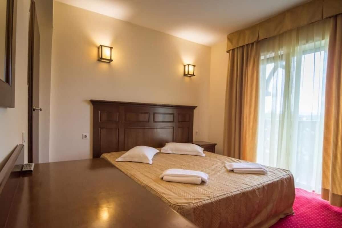 Quarto de hotel com uma cama grande, duas luminárias de cabeceira, uma cabeceira de madeira e uma janela com cortinas.