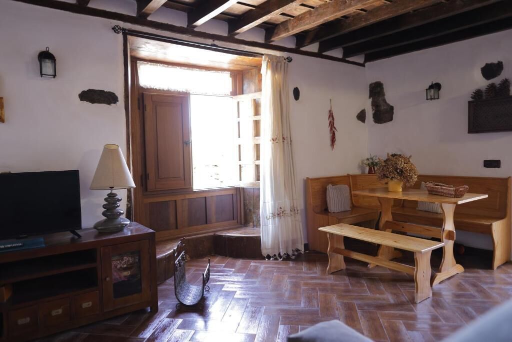 Um quarto com um banco de madeira, uma televisão sobre um móvel, uma luminária e uma janela com cortinas.