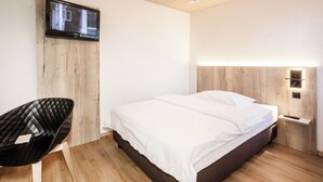 Quarto de hotel moderno com uma cama, televisão de tela plana presa a um painel de madeira, mesa de cabeceira com abajur e uma cadeira preta.