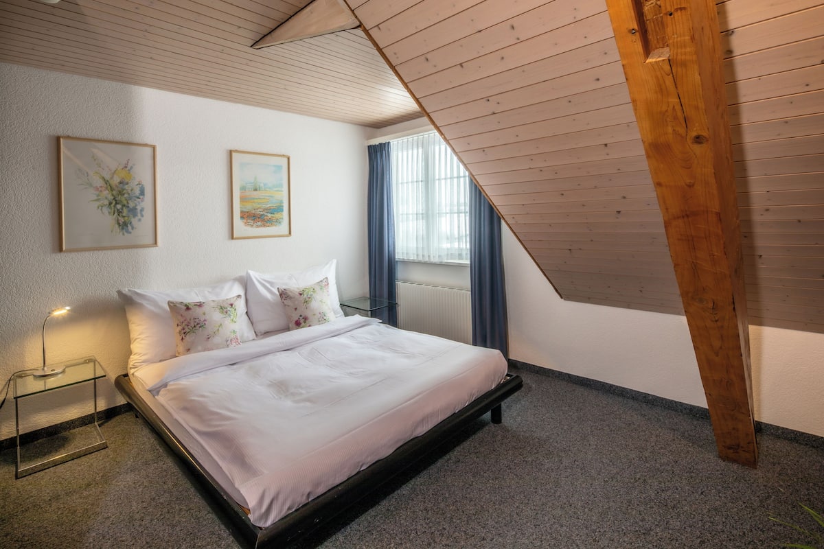 Quarto com cama, mesa de cabeceira, janela com cortinas e vigas de madeira no teto.