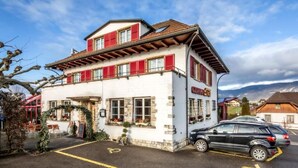 Um edifício de dois andares com fachada branca e persianas vermelhas, um estacionamento e montanhas ao fundo.