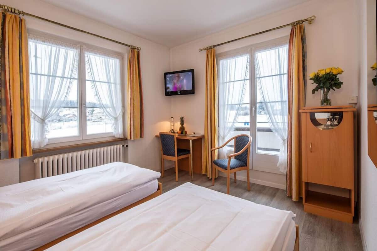 Quarto de hotel com duas camas, uma escrivaninha, uma cadeira, uma televisão e um guarda-roupa.