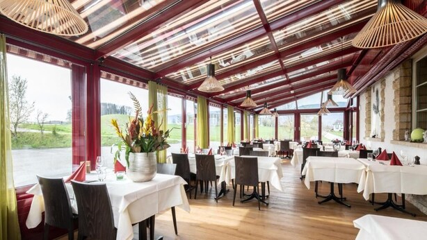 Restaurante com mesas montadas para refeições, grandes janelas e luminárias pendentes.