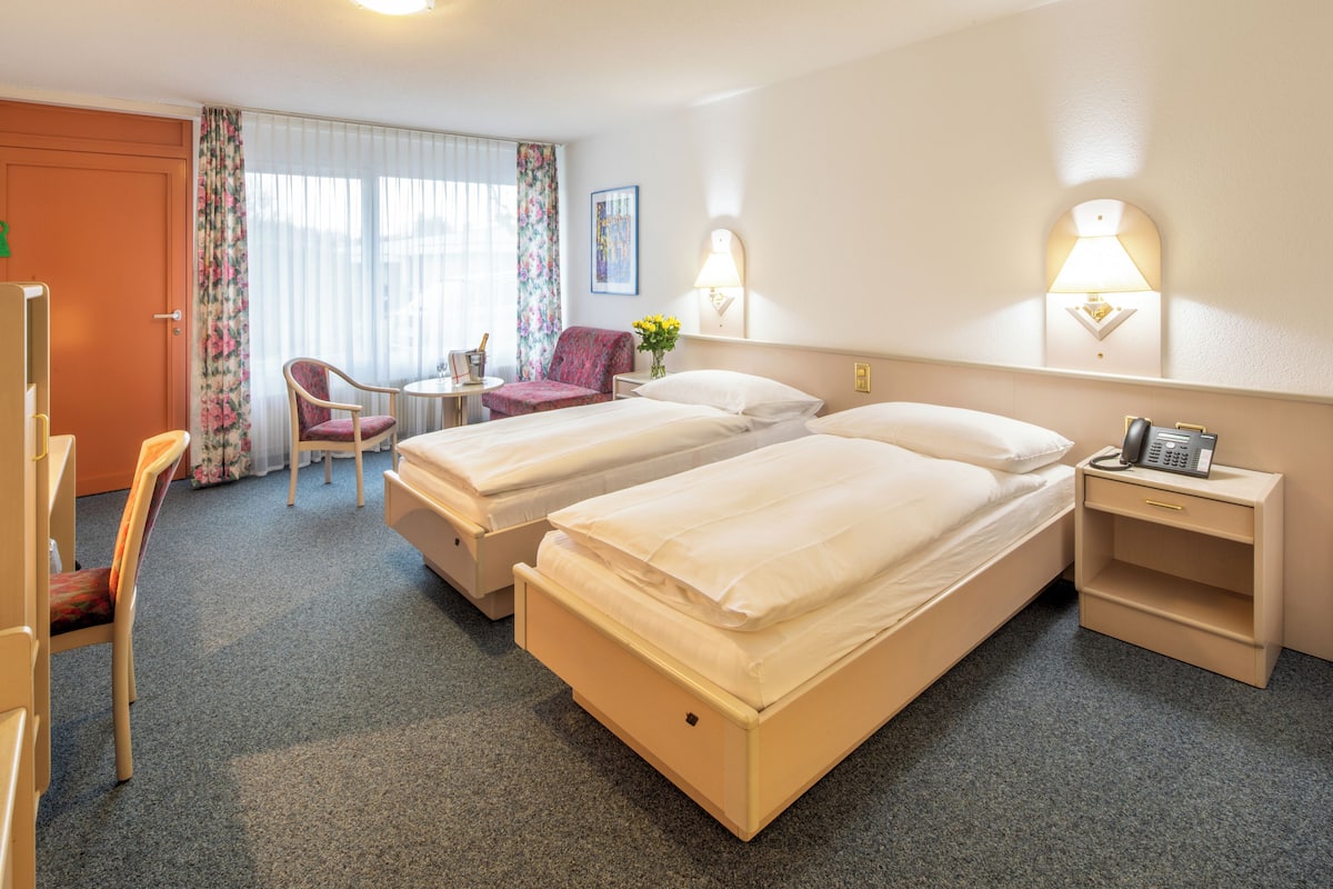 Quarto de hotel com duas camas, uma escrivaninha, uma cadeira, mesa de cabeceira com telefone e uma janela com cortinas.