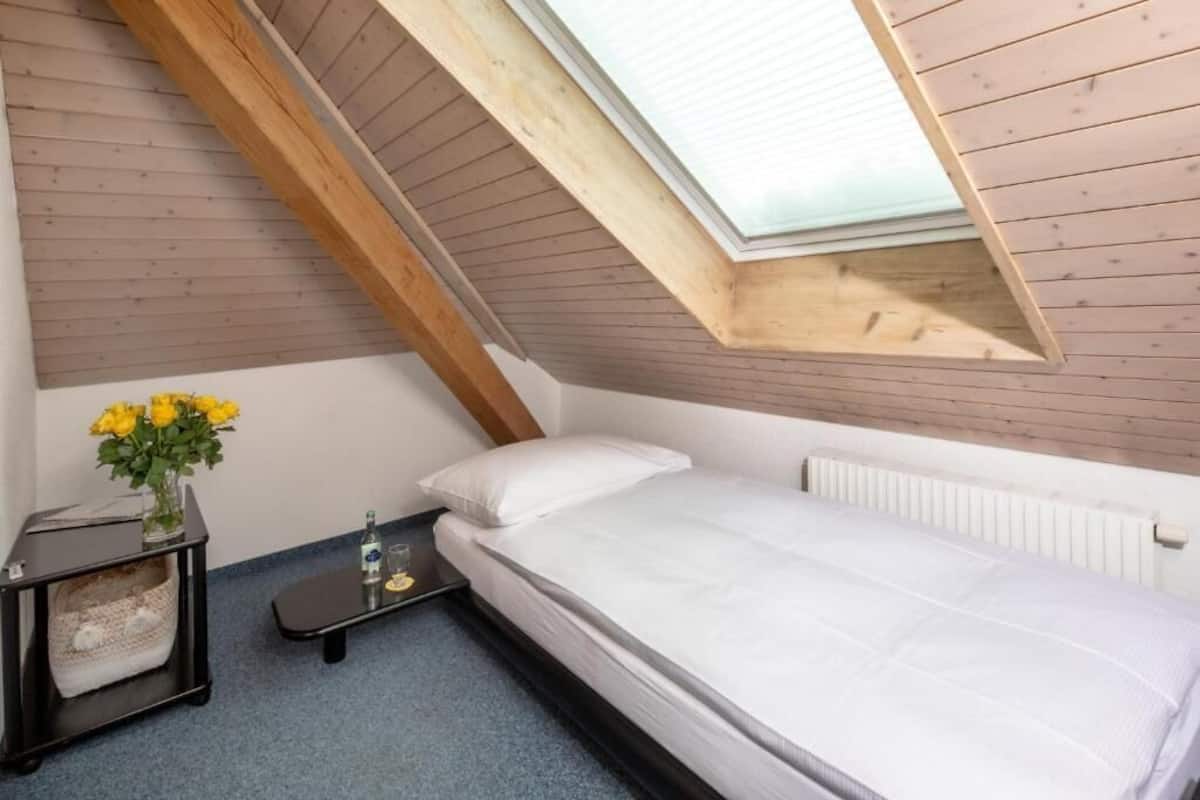Quarto com teto inclinado, uma cama, mesa de cabeceira com um vaso de flores e um radiador.
