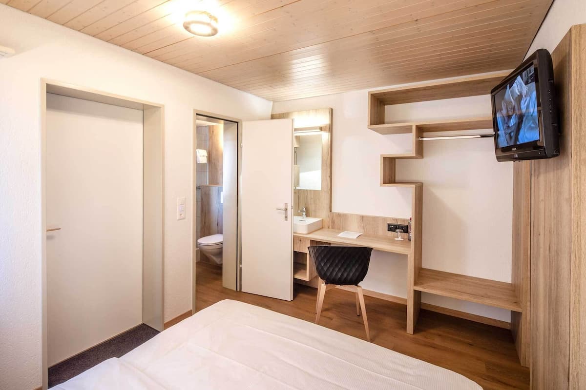 Quarto de hotel com cama, escrivaninha, cadeira, televisão e banheiro.