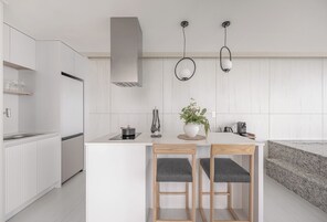 Uma cozinha moderna com armários brancos, uma ilha central com duas cadeiras, luminárias pendentes e um vaso de plantas.