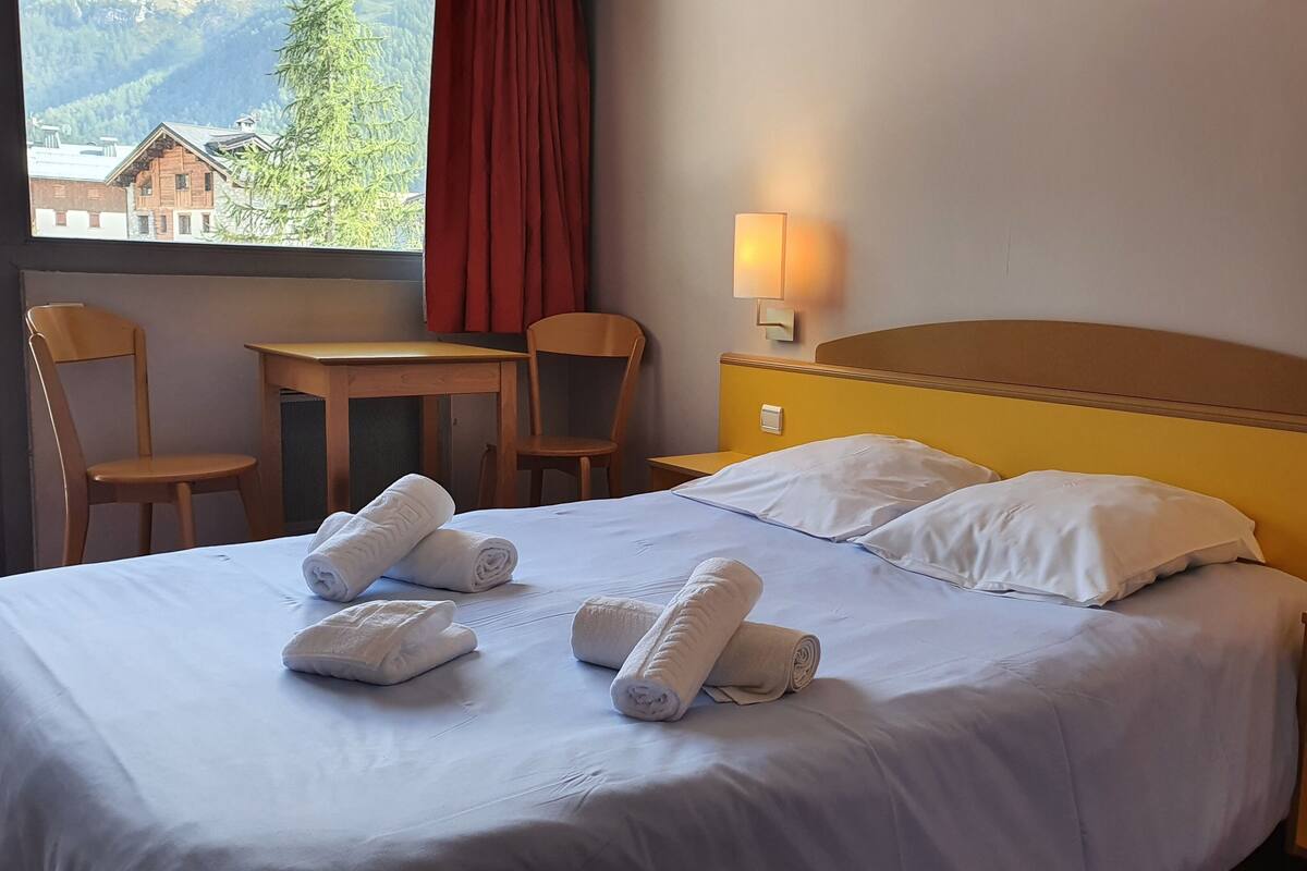 Quarto de hotel com uma cama, duas cadeiras, uma mesinha e uma janela com vista para uma montanha.
