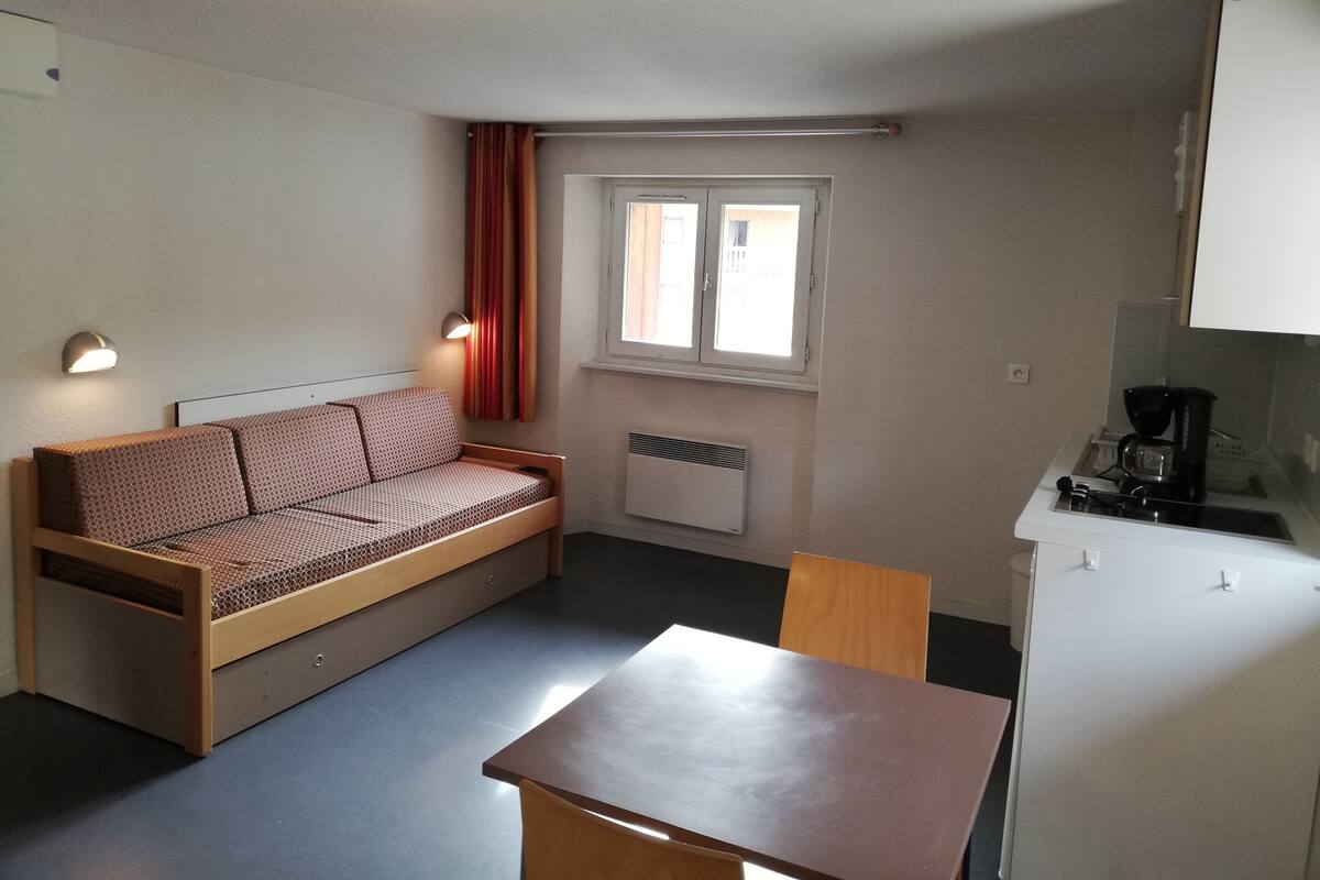 Quarto pequeno e acolhedor com sofá, mesinha, janela com cortinas, radiador e máquina de café.