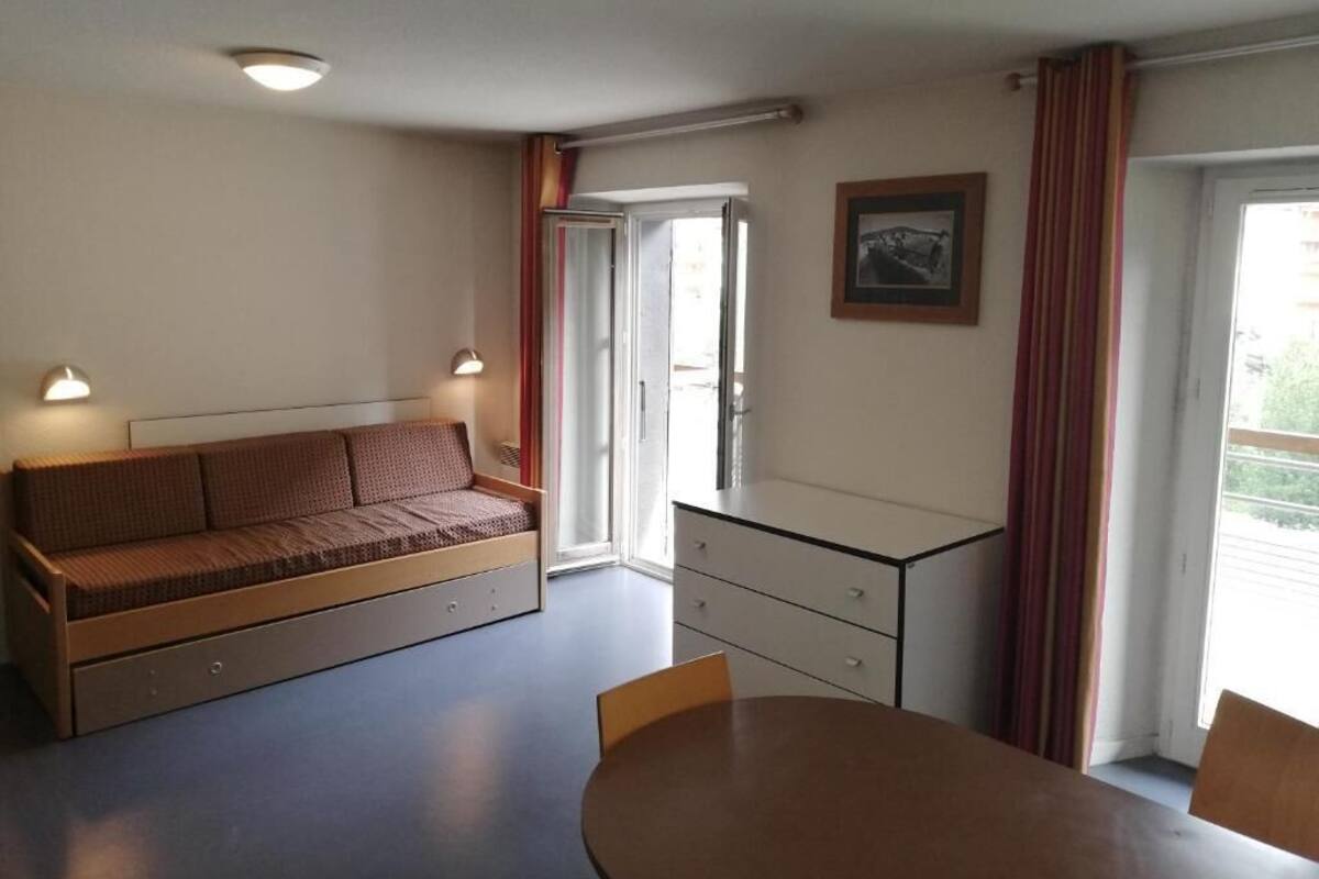 Quarto pequeno e acolhedor com sofá, uma mesinha e uma cadeira. Possui janela com cortinas, uma porta e um quadro na parede.