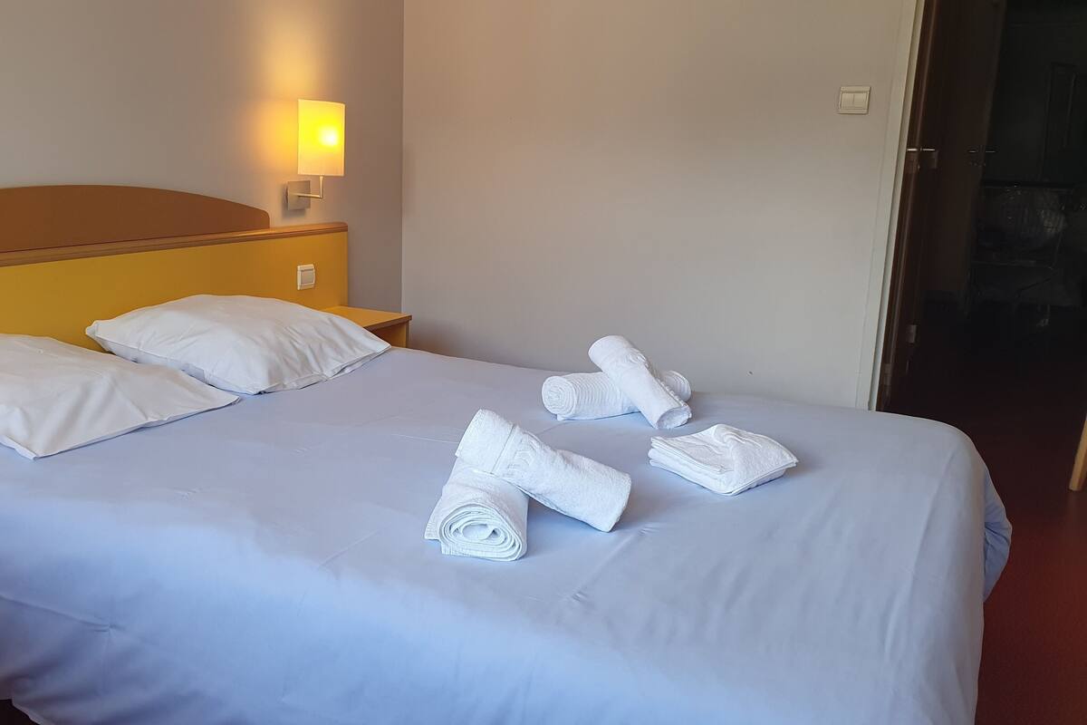 Uma cama bem arrumada com lençóis e almofadas brancos, um abajur e toalhas dobradas sobre a cama.