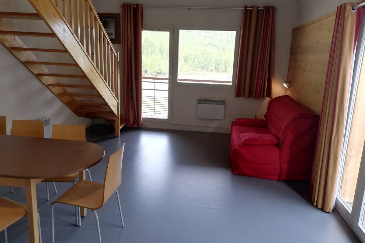 Quarto com escada de madeira, sofá vermelho, mesa de jantar com cadeiras e janela com cortinas.