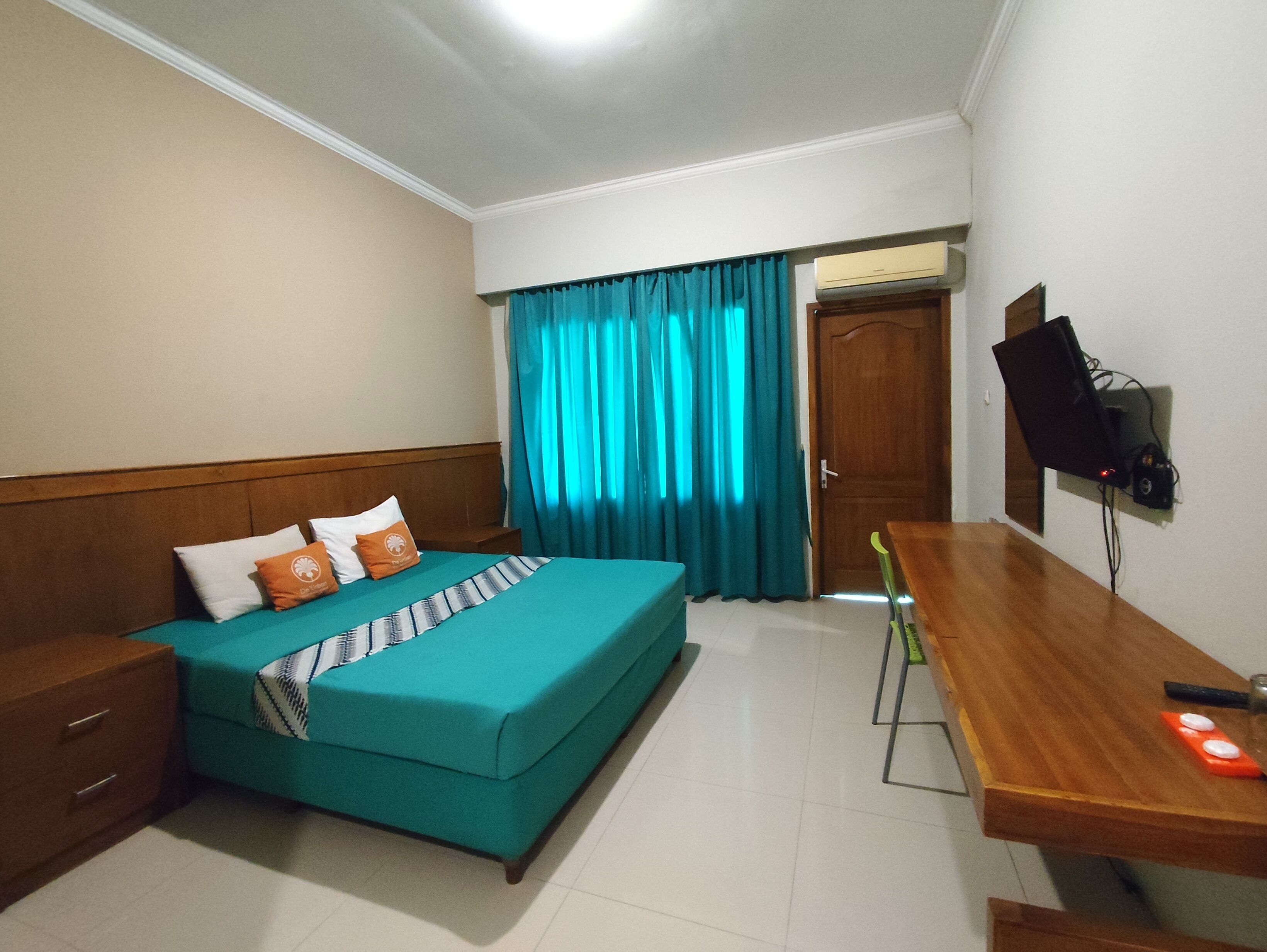 Quarto com cama, mesa de cabeceira de madeira, escrivaninha com cadeira, televisão fixada na parede e porta para outro cômodo.