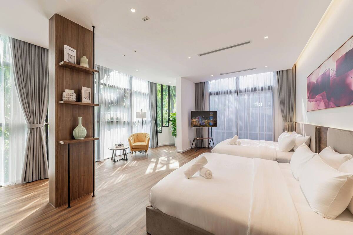 Quarto de hotel moderno com duas camas, uma estante de madeira, uma televisão e grandes janelas com cortinas.