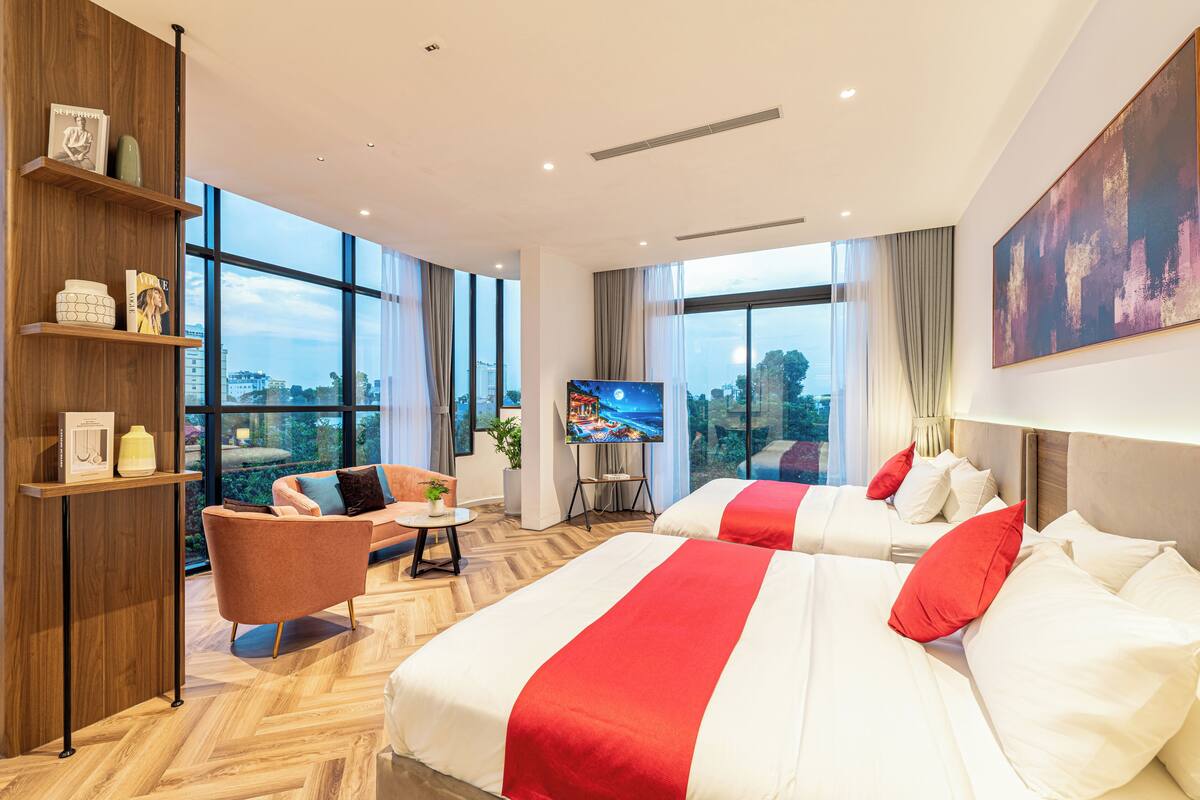 Quarto de hotel moderno com janelas amplas, TV de tela plana, sofá e duas camas com roupa de cama vermelha e branca.