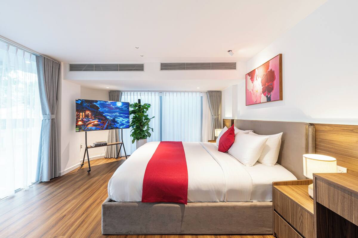 Quarto de hotel moderno com uma cama grande, televisão de tela plana, mesa de cabeceira e janela com cortinas.