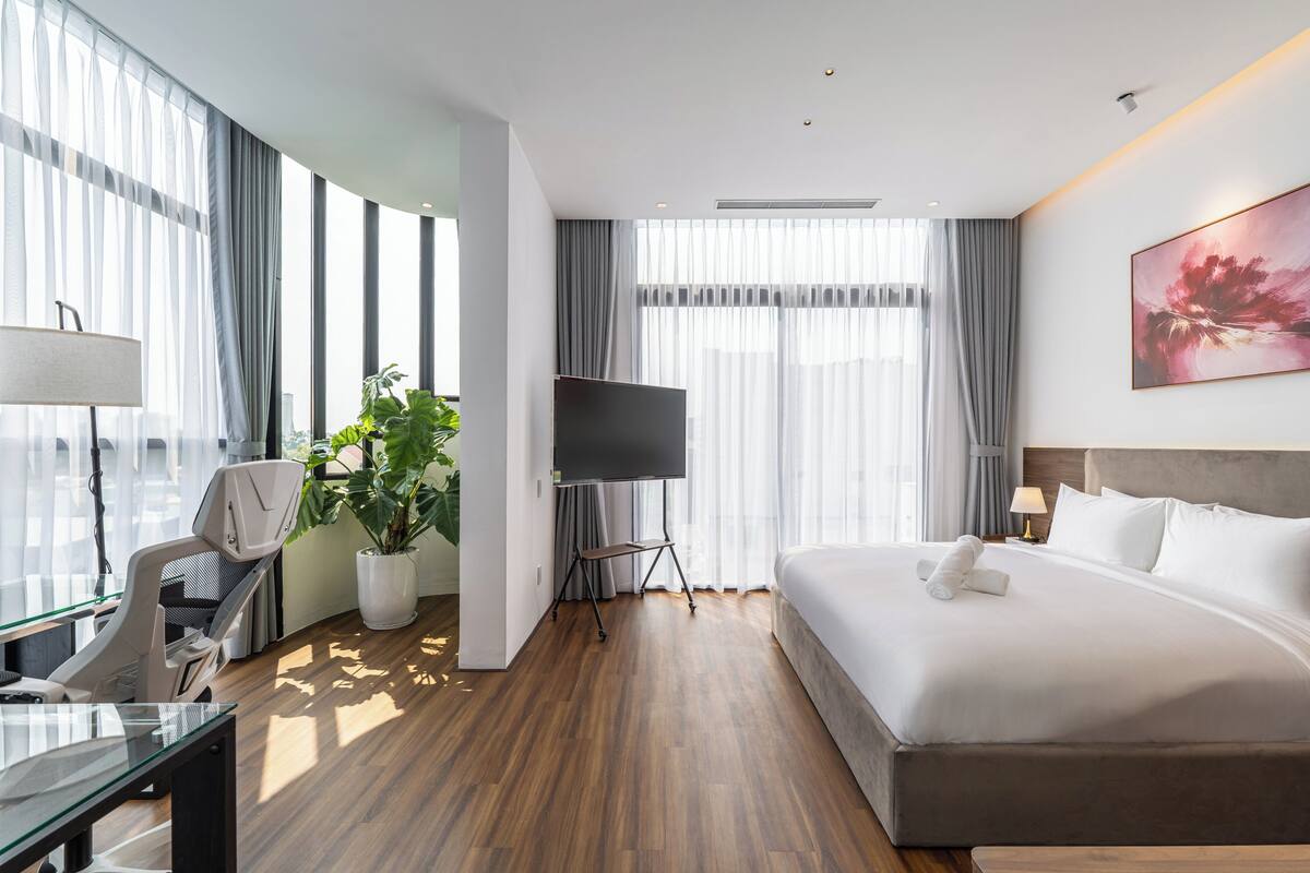 Quarto de hotel moderno com uma cama grande, uma escrivaninha com cadeira, televisão de tela plana e um vaso com planta.