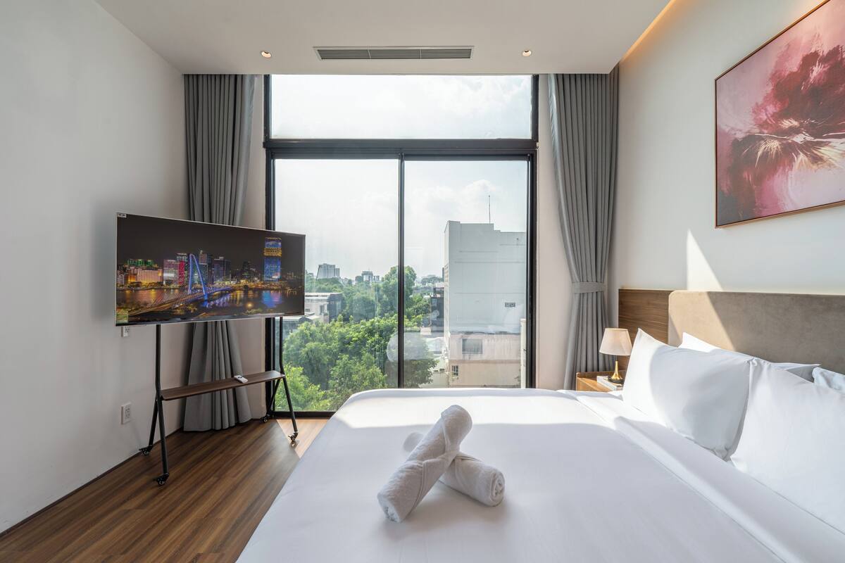 Quarto de hotel com uma cama grande, uma televisão de tela plana em um suporte, vista da cidade por grandes janelas e um quadro na parede.
