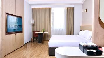 Quarto de hotel com cama, televisão, escrivaninha, cadeira e janela com cortinas.