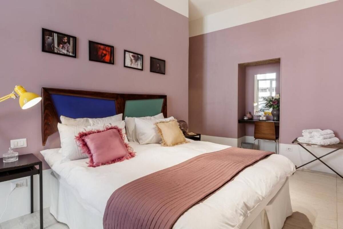 Quarto com cama, mesas de cabeceira, abajur, espelho e quadros nas paredes.