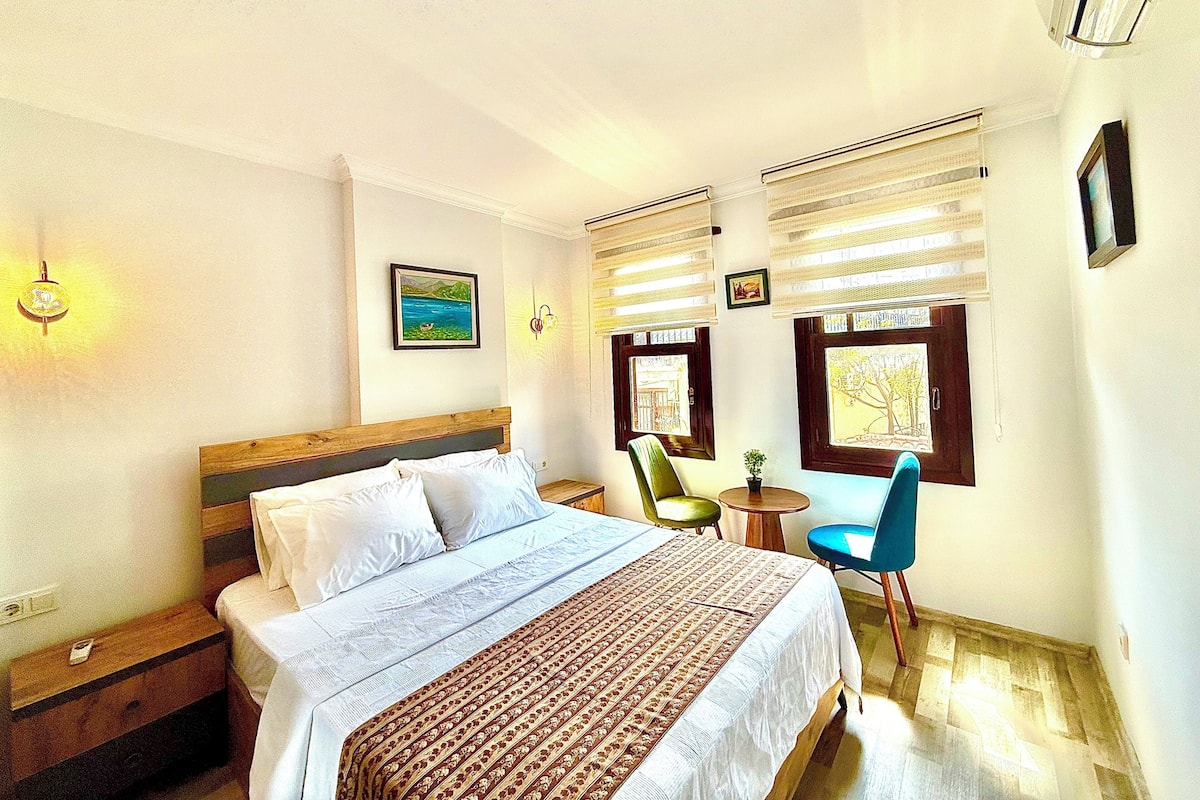 Quarto de hotel com cama, mesa de cabeceira, cadeira, mesinha e janela com persianas.