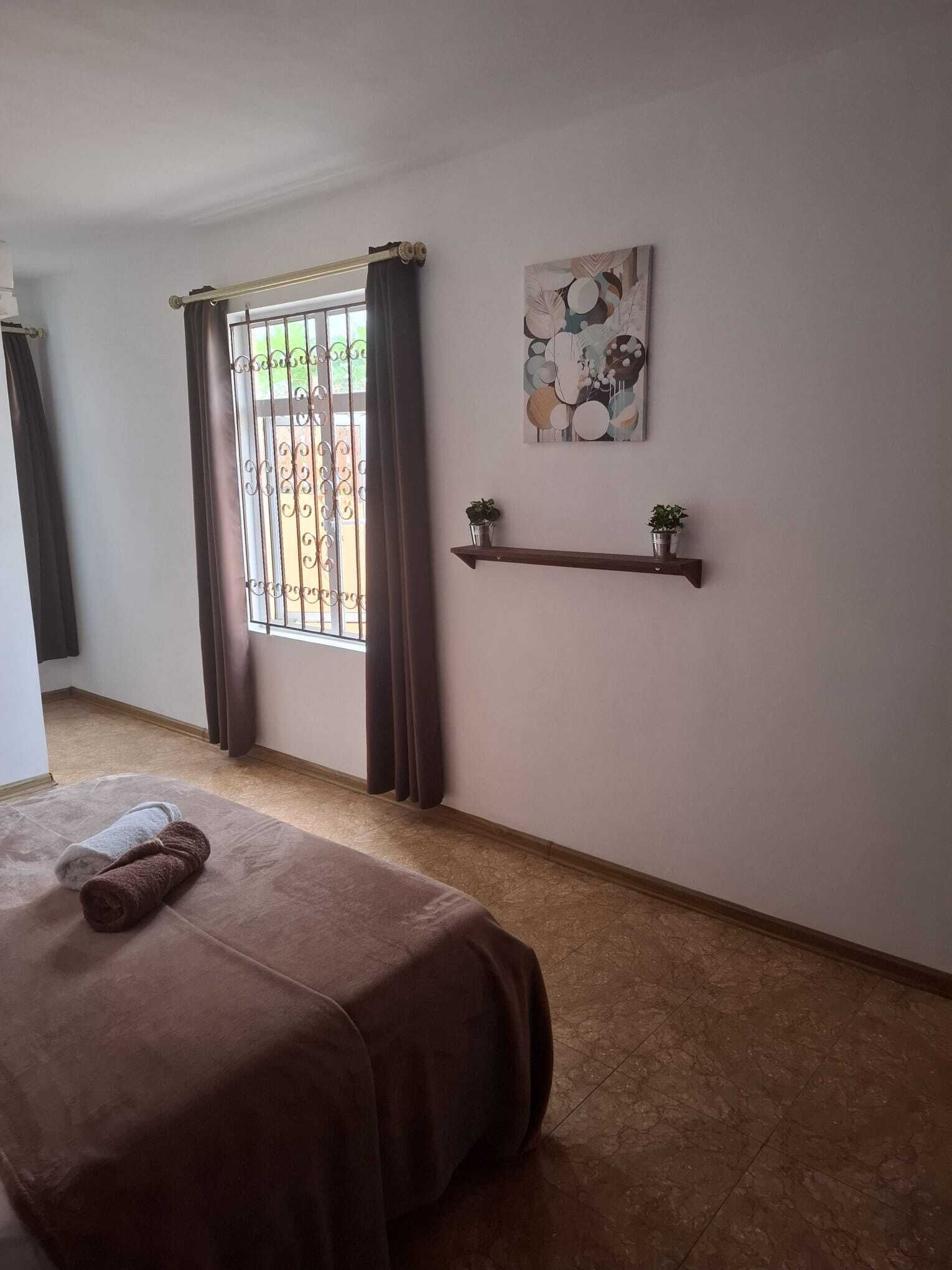 Um quarto com cama, janela com cortinas, prateleira com plantas na parede e um quadro.