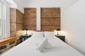 Quarto de hotel com parede de tijolos, uma cama com roupa de cama branca, duas mesas de cabeceira com abajures e uma janela.