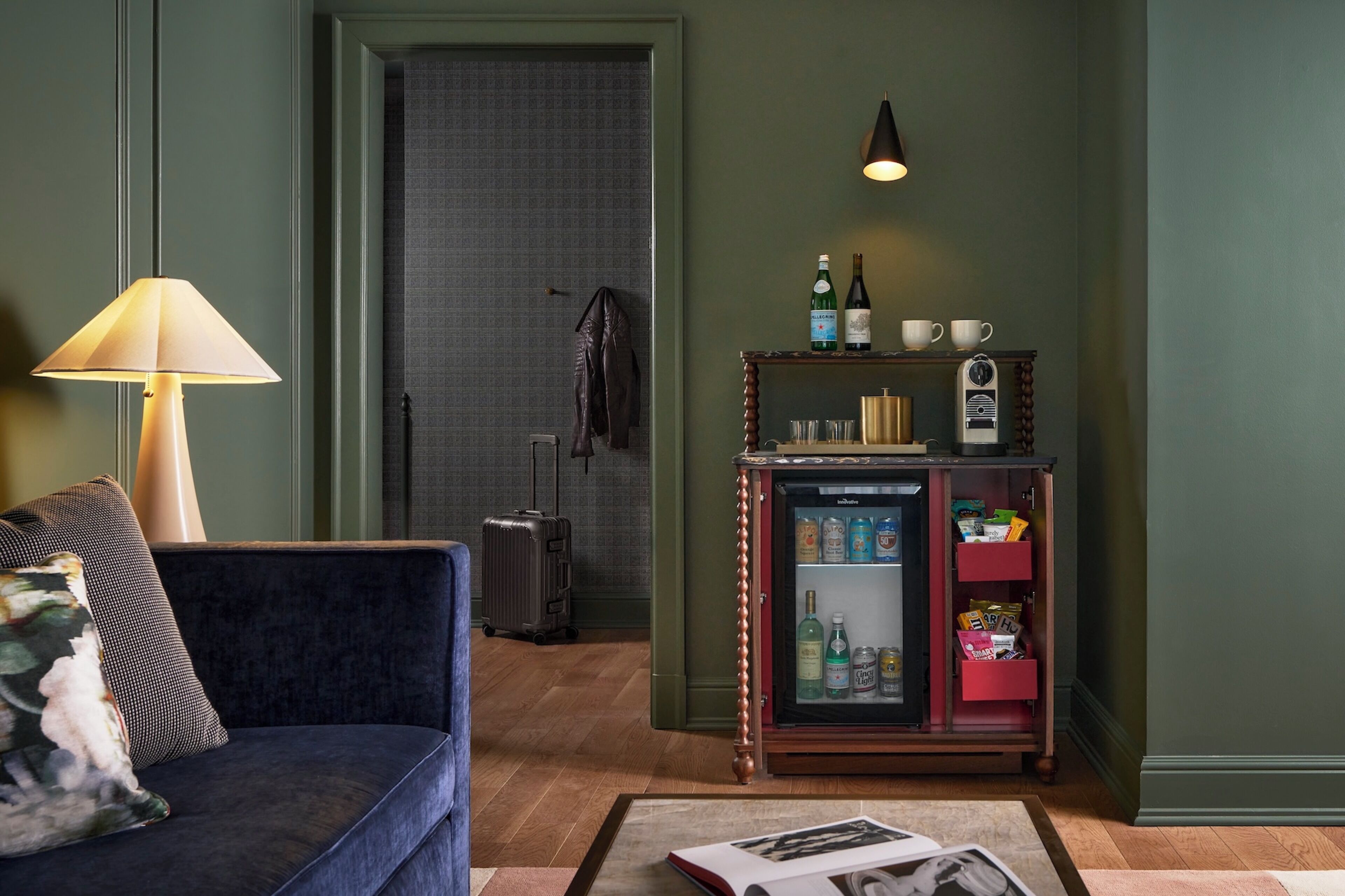 Quarto com um sofá azul escuro, uma luminária, um minibiscoitos e uma porta que dá para outro espaço.
