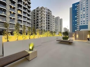 Uma área residencial urbana moderna, com edifícios altos, um espaço ao ar livre com telhado plano, assentos e plantas, e um banco de madeira.