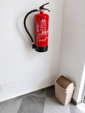 Extintor de incêndio vermelho fixado na parede com uma mangueira, lixeira bege e chão revestido com azulejos.