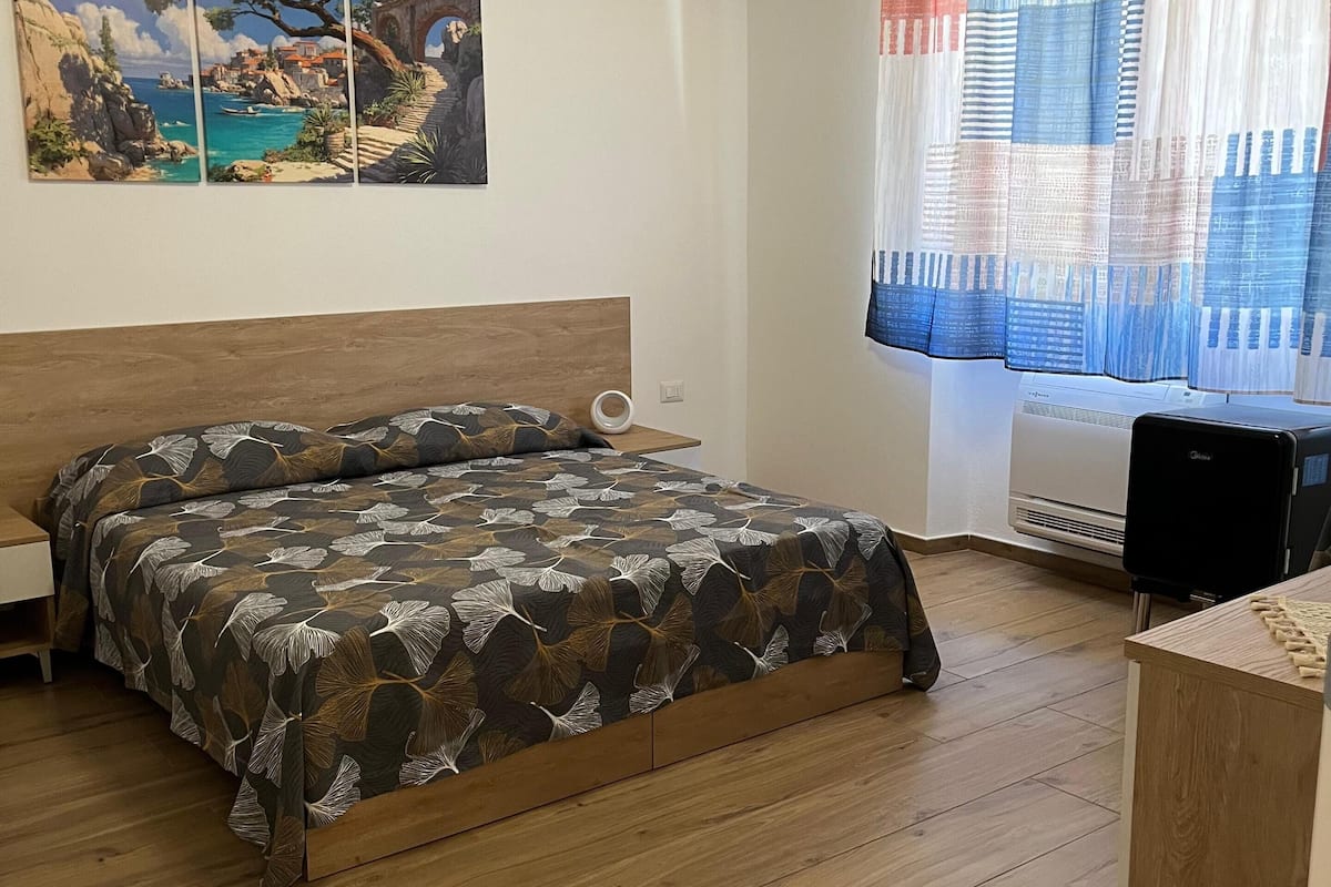 Quarto com cama, escrivaninha, janela com cortinas e ar-condicionado fixado na parede.