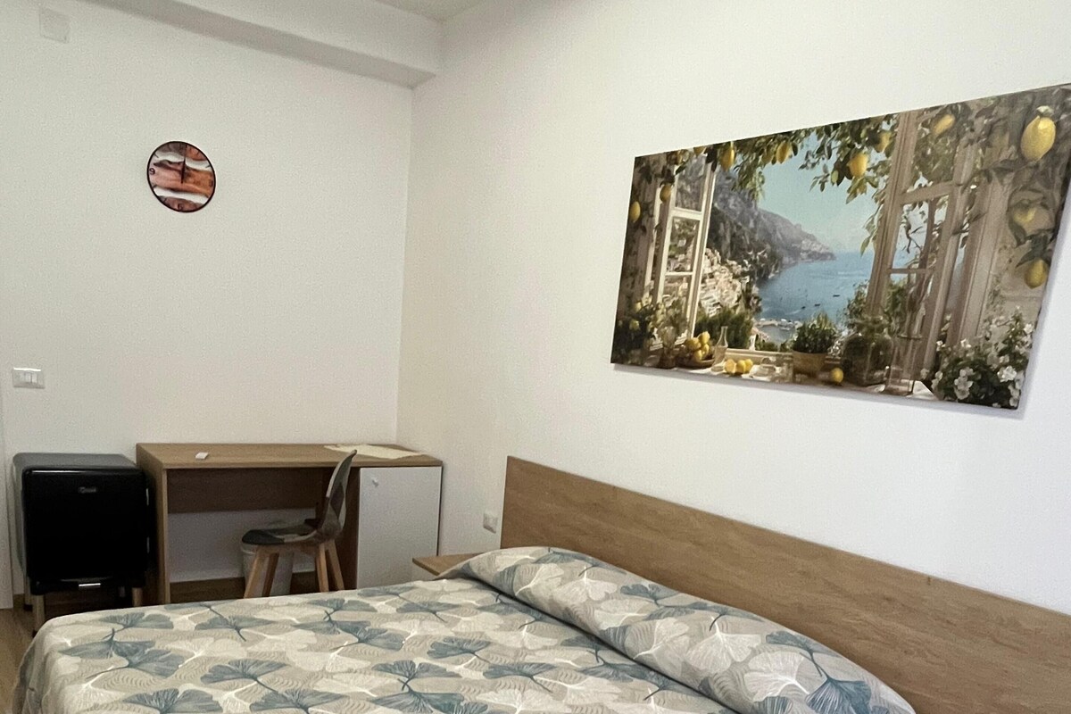 Quarto com cama, escrivaninha, cadeira, pequeno armário e quadro na parede.