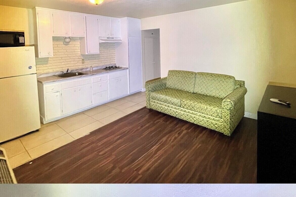 Quarto com cozinha compacta, sofá verde com padrão, cama com colcha, escrivaninha com cadeira e porta vermelha.