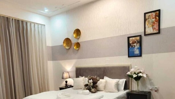 Quarto de hotel com uma cama, uma mesinha de cabeceira, um abajur, um urso de pelúcia, um vaso com flores e um quadro na parede.