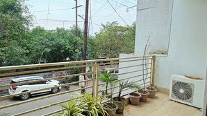 Varanda com plantas em vasos, um carro branco estacionado na rua e um ventilador no chão.