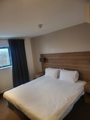 Quarto de hotel com uma cama grande, cabeceira, mesa de cabeceira, luminária e janela com cortinas.