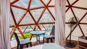 Quarto com uma janela grande, uma cama, duas cadeiras, uma mesinha e um ventilador.