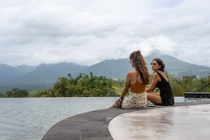 Duas pessoas sentadas à beira de uma piscina, com uma paisagem montanhosa ao fundo.