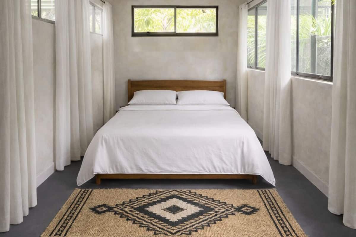 Um quarto com uma cama, ventilador de teto, janelas amplas e um tapete estampado.