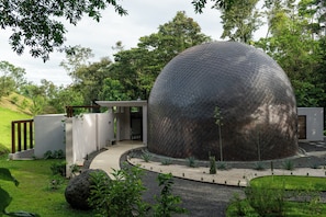 Um edifício esférico com exterior texturizado, rodeado por um jardim paisagístico com caminhos e cactos.