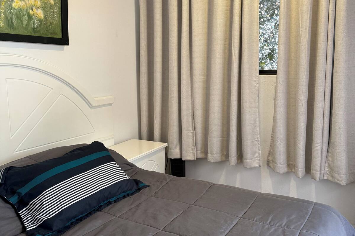 Quarto com cama, mesa de cabeceira, um quadro e janela com cortinas.